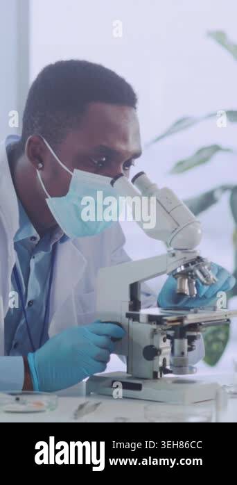 Black scientist examines slide under microscope, wearing surgical mask ...