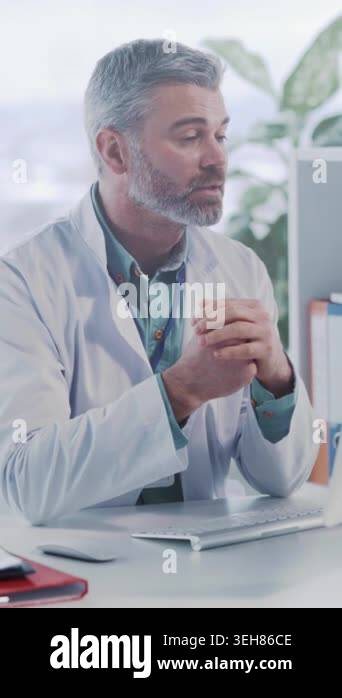 white male doctor seated at desk, contemplative expression and clasped ...