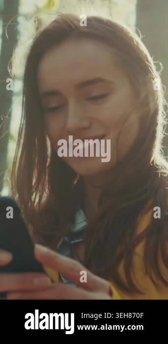 caucasian woman smiling under sunlit trees, checking smartphone screen ...