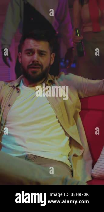 skeptical man on red couch reacting to lively neon party, arms ...