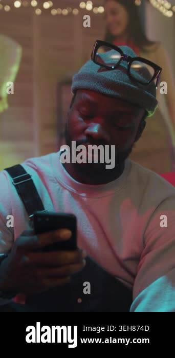 black man checking phone at house party, beanie with glasses, overalls ...