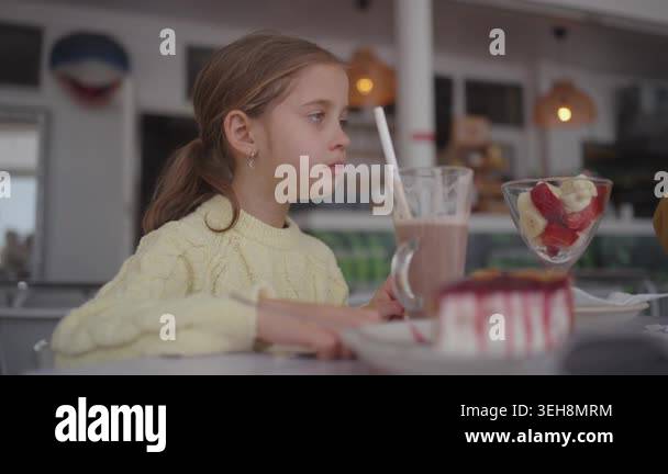 Little girl sits at cafe table looking at her dessert. She has ice ...