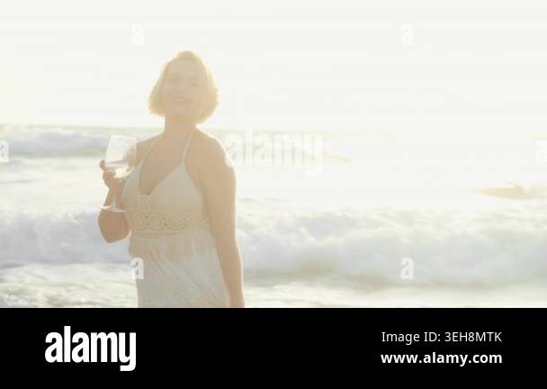 Woman holding wine glass walking along rocky beach with ocean waves and ...