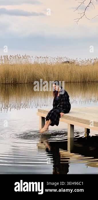 woman sitting on dock dipping feet into lake water, plaid robe, with ...