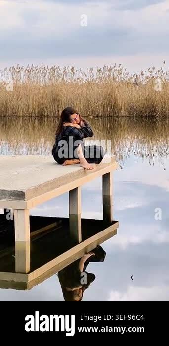 woman sitting on concrete dock near lake, barefoot, reflecting on calm ...