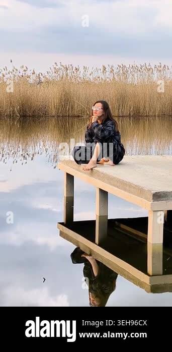 young woman on pier, reflecting on calm water with distant reeds and ...