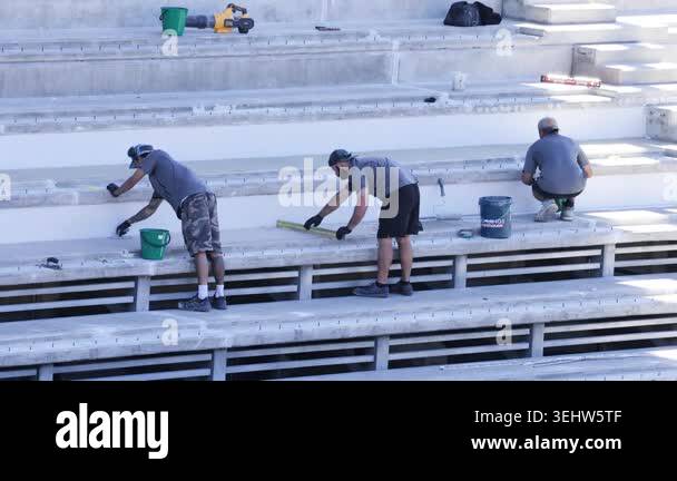 Three workers painting benches in a large outdoor stadium under bright ...