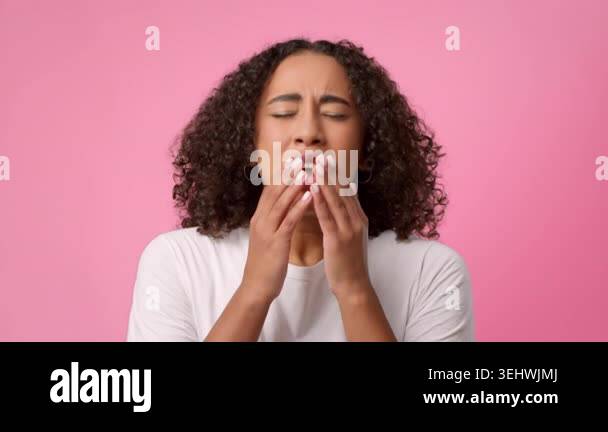 A woman with curly hair stands in front of a pink background. She has ...