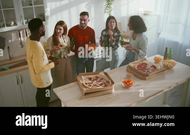 A group of friends stands around a table in a sunlit kitchen sharing ...