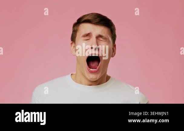A young man with light skin yawns widely in front of a pink background ...