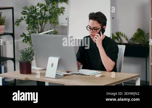 A man with light skin and glasses is engaged in a phone call while ...