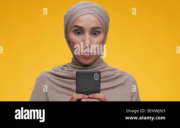 A woman wearing a hijab holds her smartphone, focusing on the screen ...