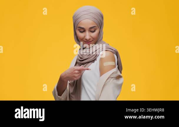 A woman in a headscarf points at a bandage on her arm. She stands in a ...