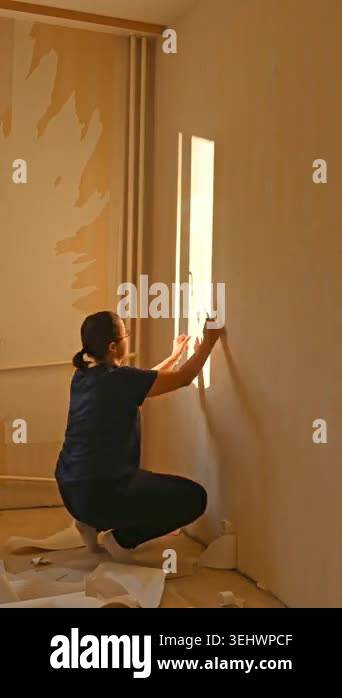 Woman scraping peeling wallpaper from interior corner wall at sunset in ...