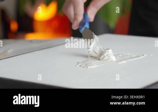 A hand spreads white texture paste on a canvas or panel with a small ...