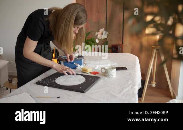 Woman applies white texture paste on black canvas at covered table with ...