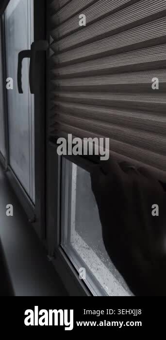 Hand lifts pleated window blinds, revealing a rainy city view through a ...