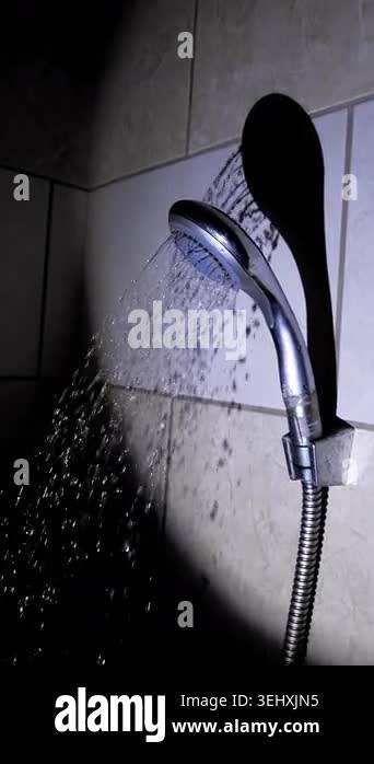 A handheld shower head sprays water in a dark, tiled bathroom stall. A ...