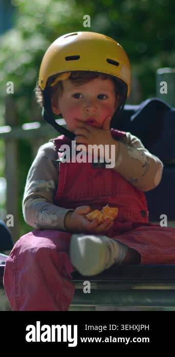 Young child in protective helmet enjoys tangerine while sitting outside ...