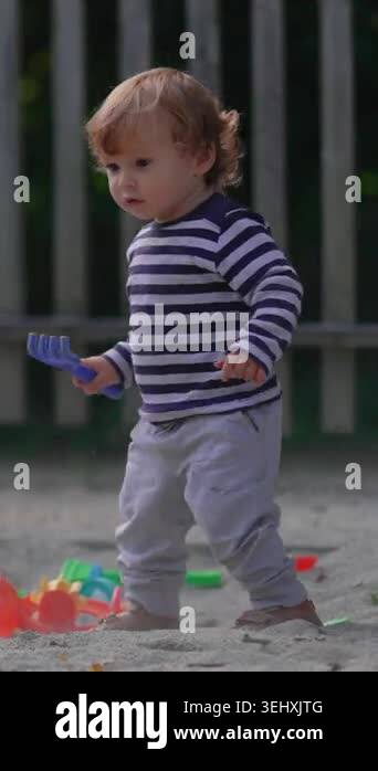 Toddler walking across sandy play area with small rake at public park ...