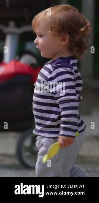 Toddler carrying small beach toys through public park sandbox capturing ...