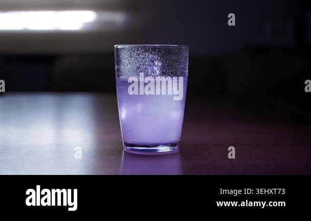 A chilled glass filled with purple liquid and ice sits on a dark table ...