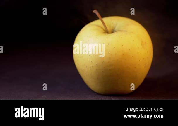 A whole yellow Golden Delicious apple sits on a dark surface surrounded ...