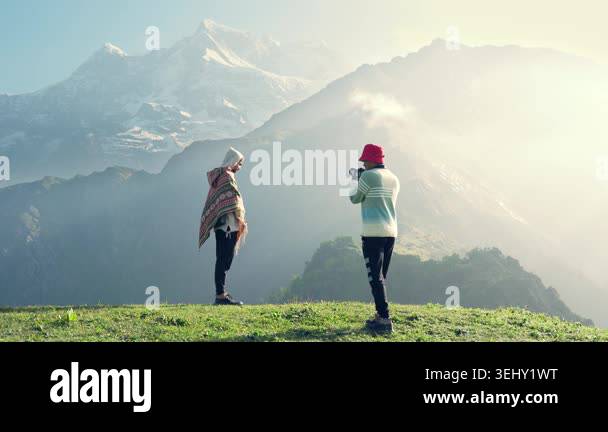 Two people stand on a green hill overlooking a vast mountain range ...