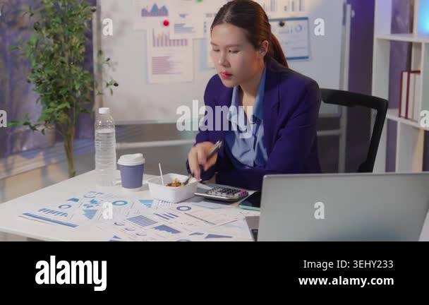 An Asian woman is working overtime and eating while sitting at a wooden ...