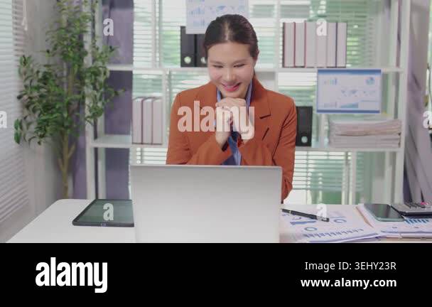 An Asian woman is working on documents on a laptop in an office. She is ...