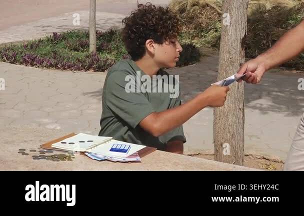 A teenage boy with curly hair sitting outdoors at a table with a ...