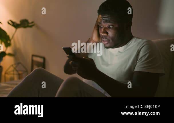 A man with dark skin sits on a couch, holding a smartphone. He looks ...