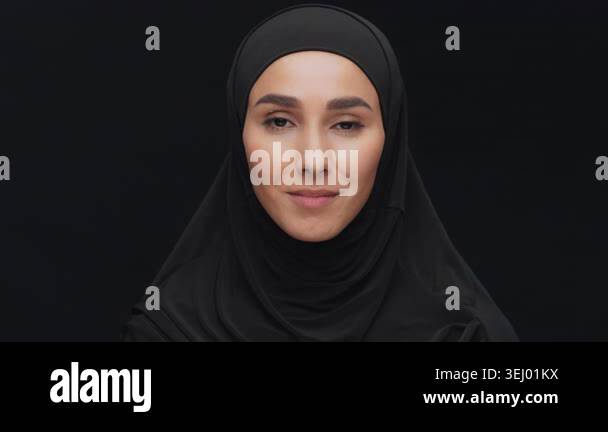 Woman dressed in a black hijab stands in front of a dark background ...