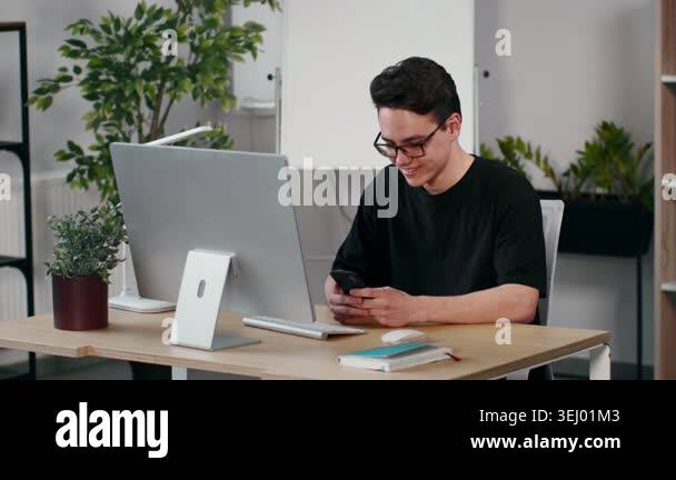 A young man with light skin and glasses smiles while using a smartphone ...
