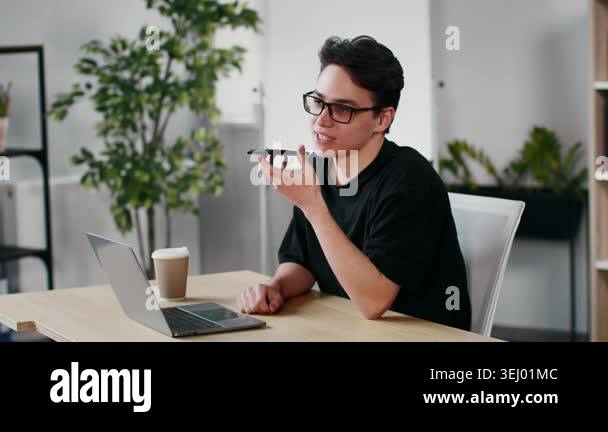 A young man with light skin and glasses is sitting at a desk in a ...