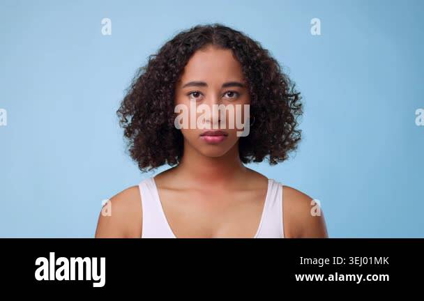 A young woman with curly hair stands confidently against a light blue ...