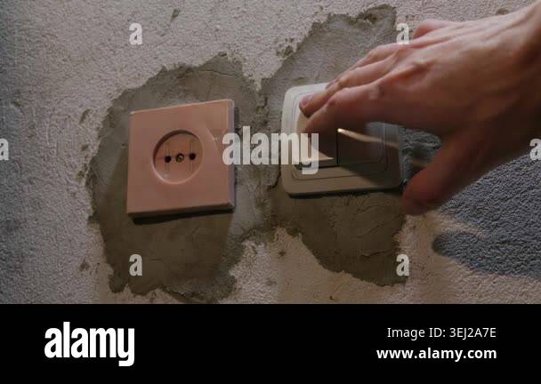 A hand presses a white light switch next to an electrical outlet on a ...