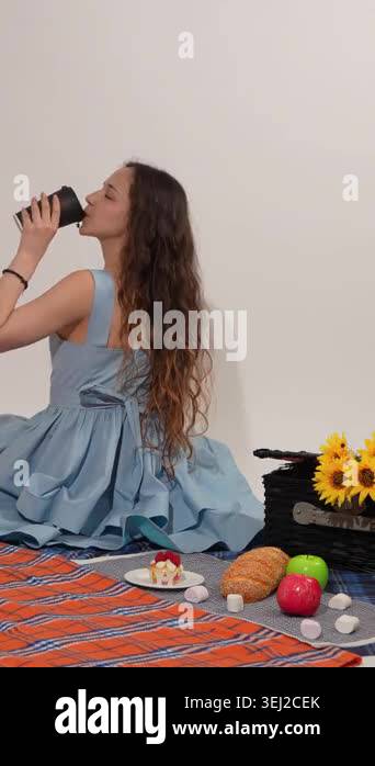 A beautiful young woman sits on a plaid blanket, drinks coffee from a ...