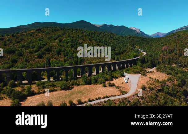 Aerial footage of a train passing over an arched stone bridge in a lush ...