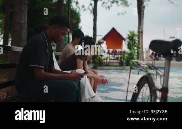 Three diverse young friends are sitting on a park bench, each engrossed ...