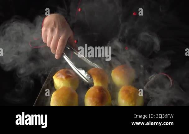 Chef in black attire with red accents lifts a golden roll using tongs ...