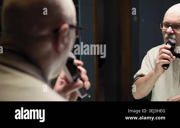 Pension man wearing glasses using a trimmer in his hands to shave the ...