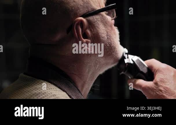 Old man wearing glasses using a trimmer in his hands to shave himself ...