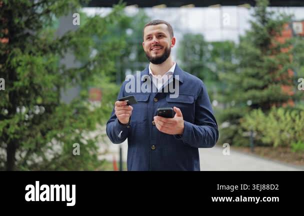Smiling bearded man in jacket holding card and smartphone, looking up ...