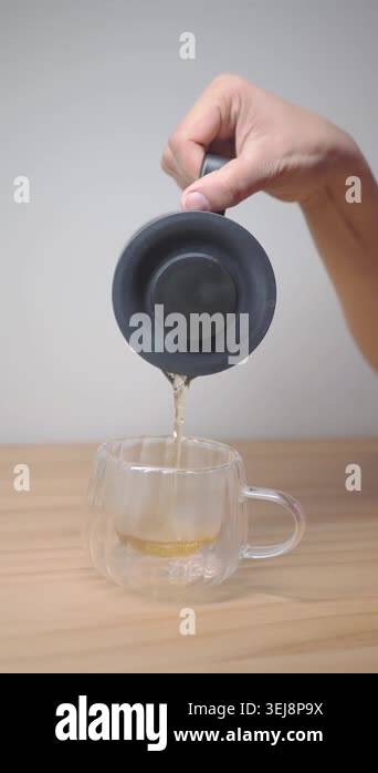 A hand pours hot tea from a pot into a clear glass cup on a wooden ...