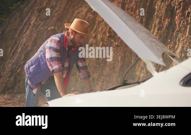 White man inspecting car engine closely beside rocky hillside, plaid ...