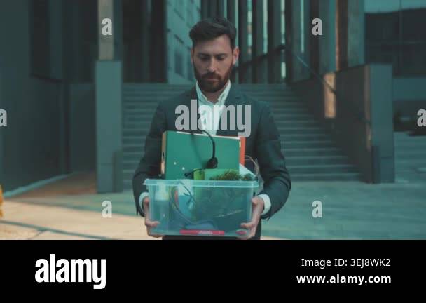 somber caucasian executive carrying box pauses on corporate plaza ...