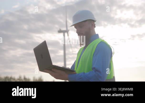 Bearded white technician checking laptop data, wind turbine in ...