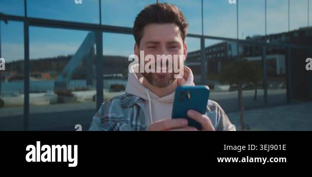 smiling caucasian man scrolling smartphone outdoors near reflective ...