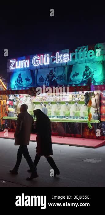 neon fair booth at night, couple in coats stroll past colorful game ...